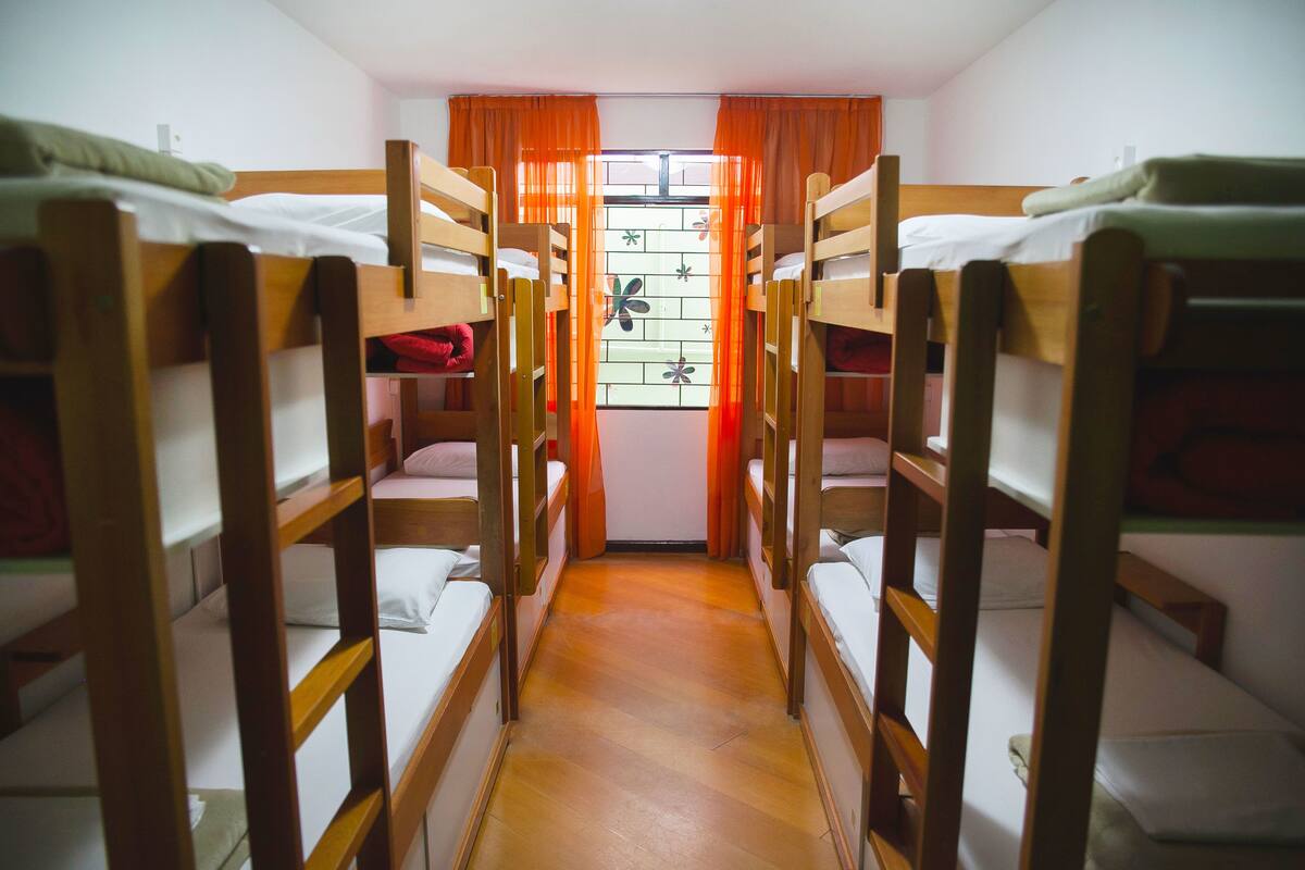 Quarto com quatro beliches alinhados, janela com cortinas laranjas e chão de madeira.