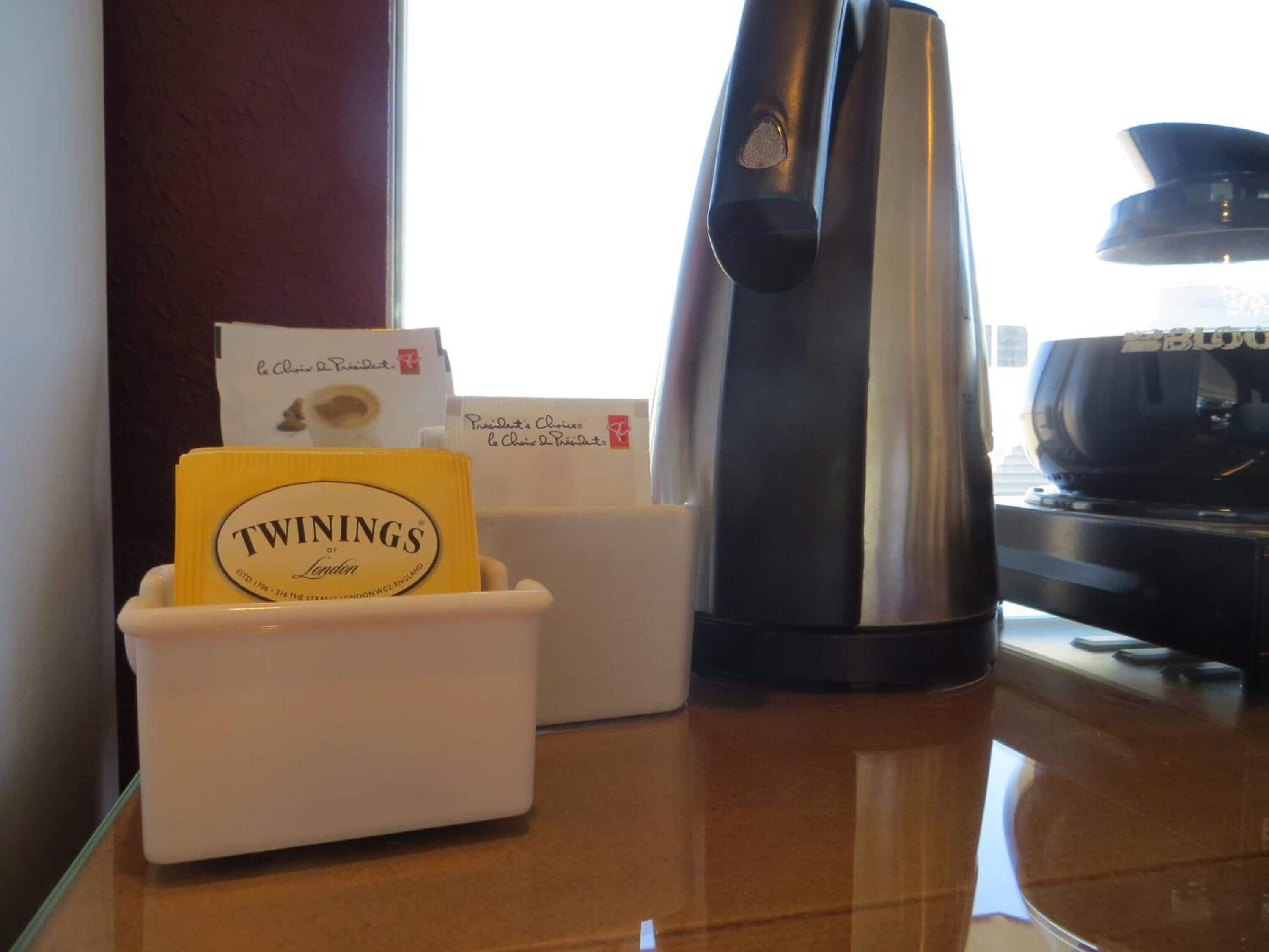 Uma cafeteira, um recipiente branco com uma caixa amarela identificada com 'TWININGS' e um pedaço de papel branco com texto sobre uma mesa.