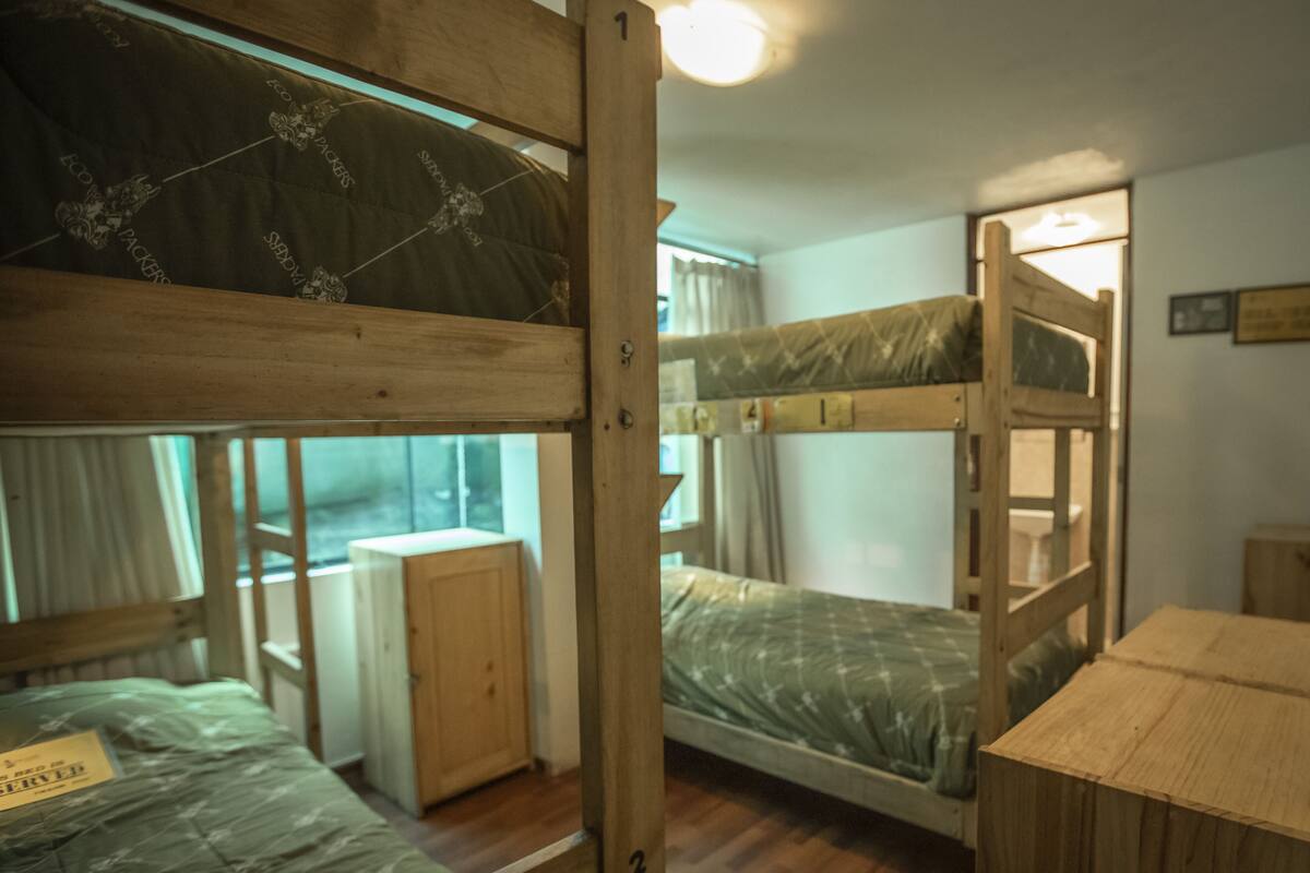 Quarto com beliches de madeira, uma escrivaninha e uma janela com cortinas.