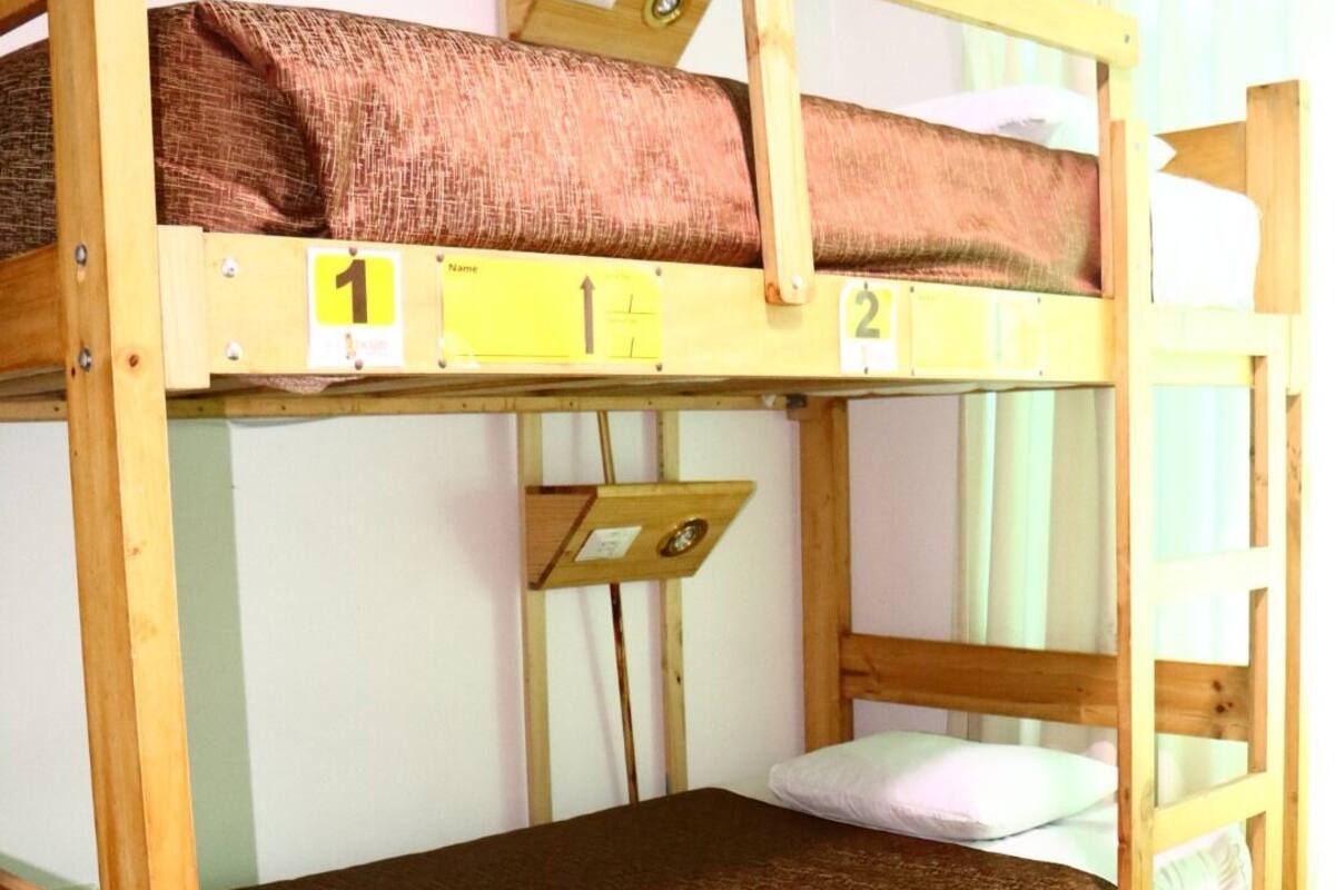 Uma cama beliche com dois níveis, cada um com colchão e travesseiro. Existem duas etiquetas numeradas na estrutura da cama, e uma escada fixada ao lado.