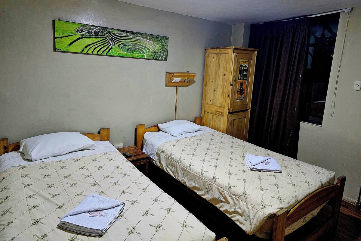 Quarto com duas camas, um guarda-roupa de madeira, um quadro na parede e uma janela com cortinas.