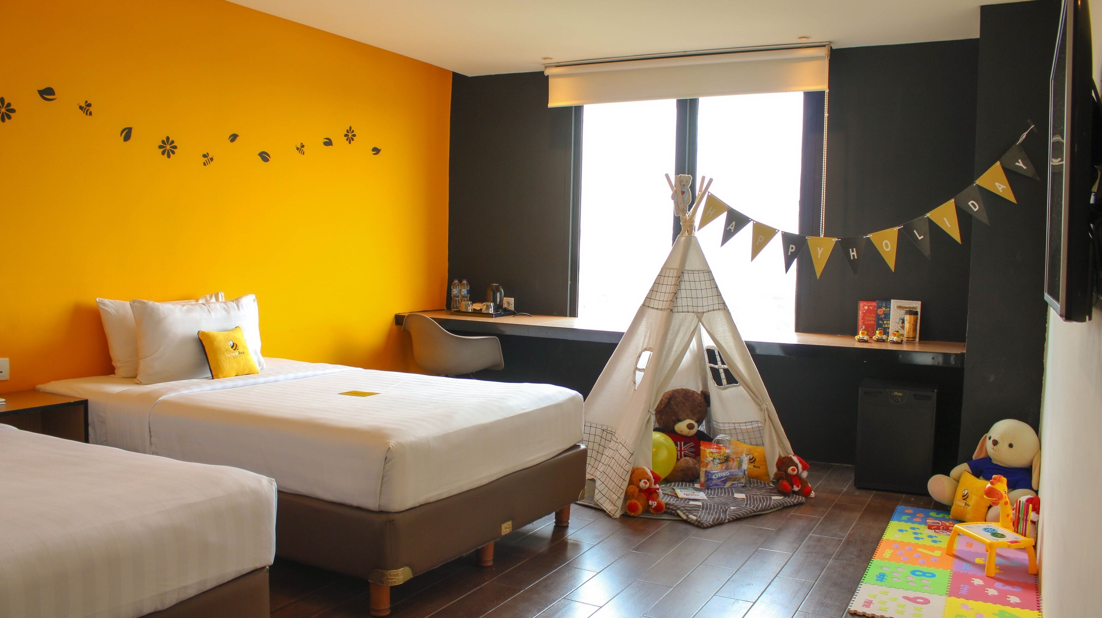 Quarto de hotel com cama, escrivaninha, cadeira, janela e uma estrutura semelhante a uma tenda com brinquedos.