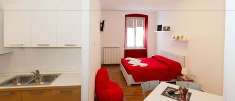 Quarto compacto com cozinha compacta, cama com roupa de cama vermelha, um pufe vermelho e uma pequena mesa com um controle remoto.