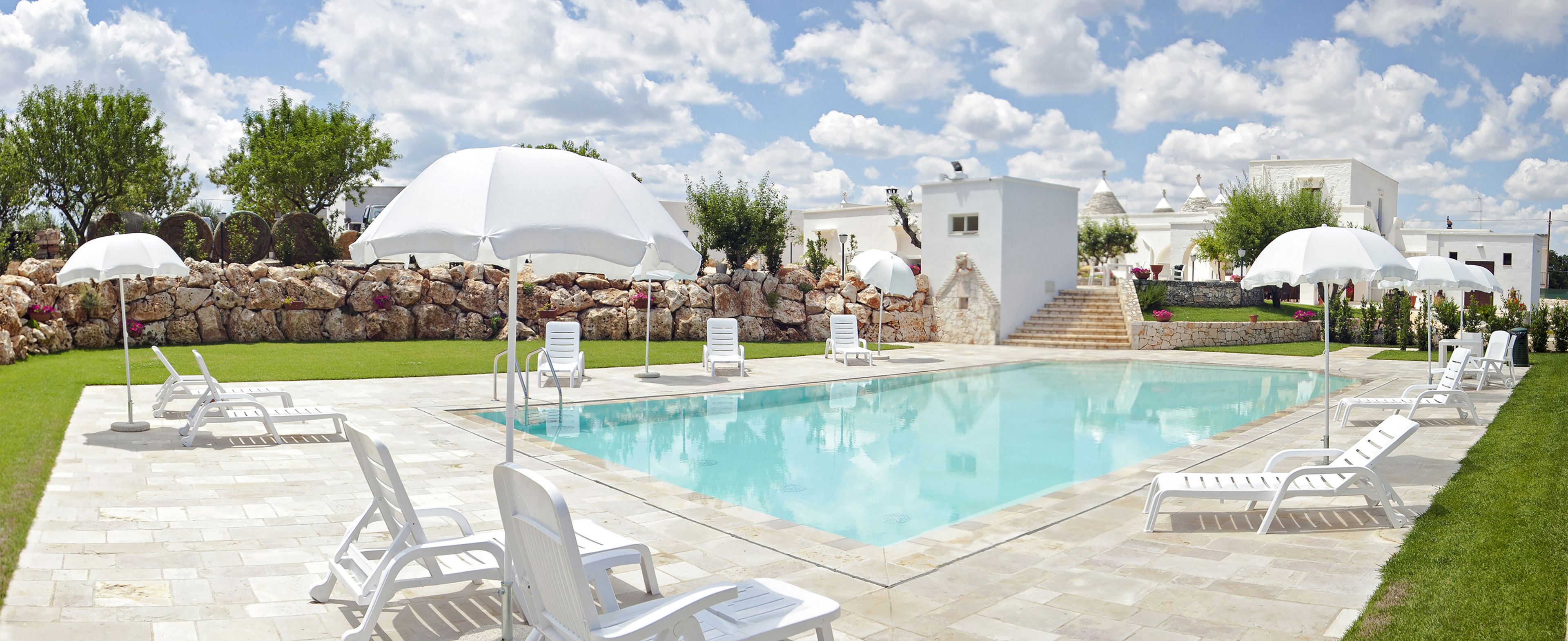 Piscina com cadeiras de descanso brancas e guarda-sóis, cercada por um muro de pedra e vegetação.