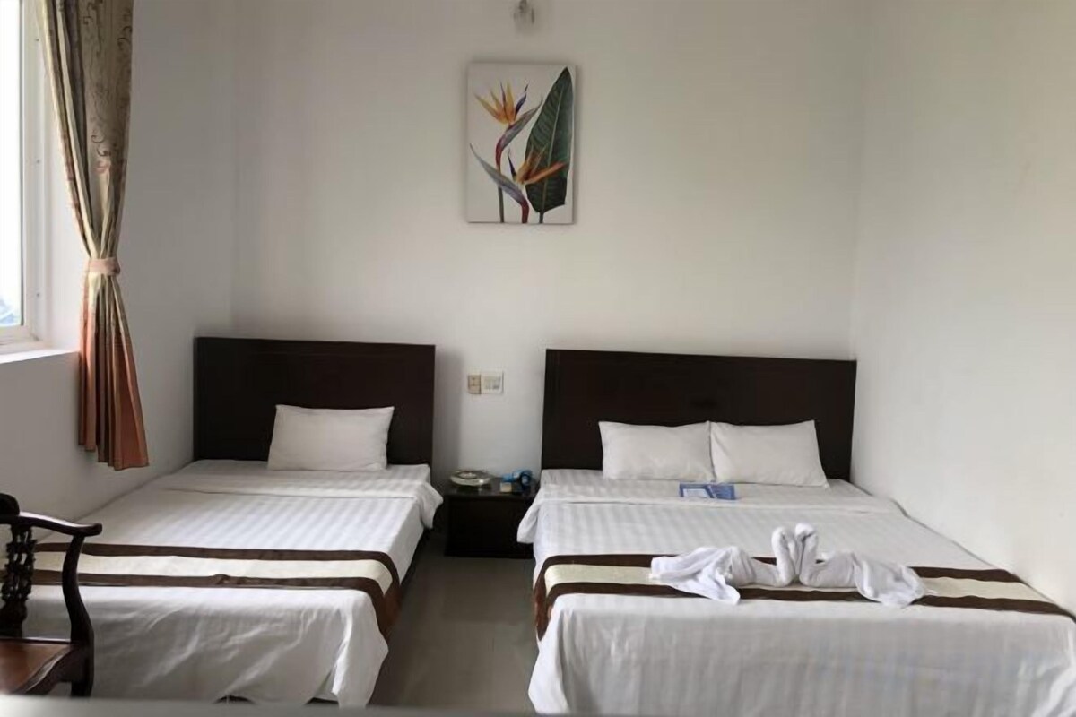 Quarto de hotel com duas camas, mesa de cabeceira, um quadro e janela com cortinas.