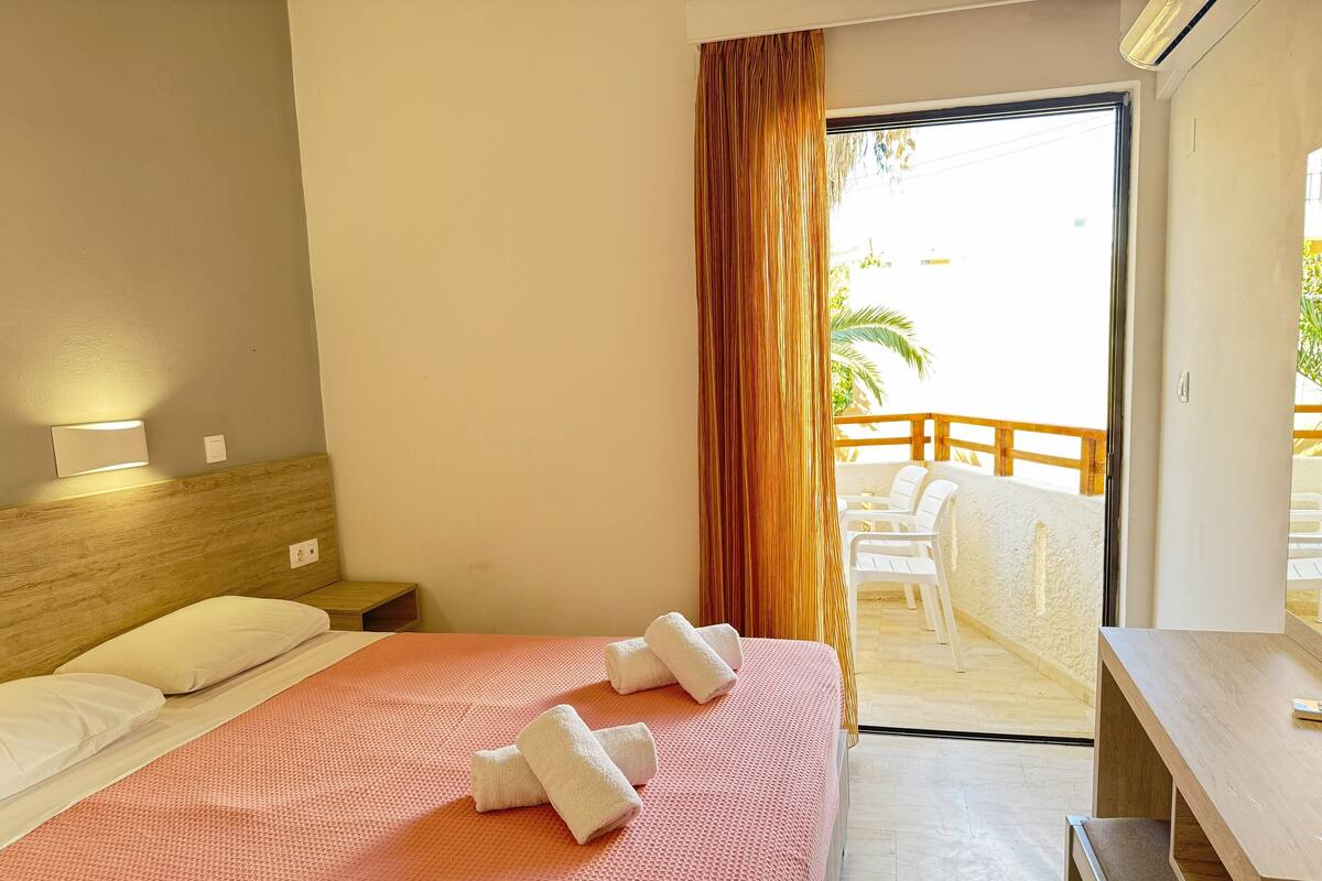 Quarto de hotel com cama, escrivaninha e varanda com mobiliário de exterior.