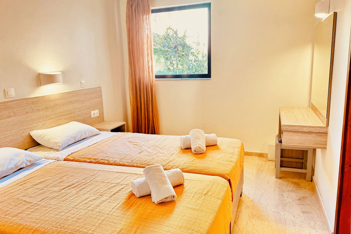 Quarto de hotel com duas camas, cabeceira de madeira, mesa de cabeceira, janela com vista para vegetação e pequena escrivaninha com espelho.