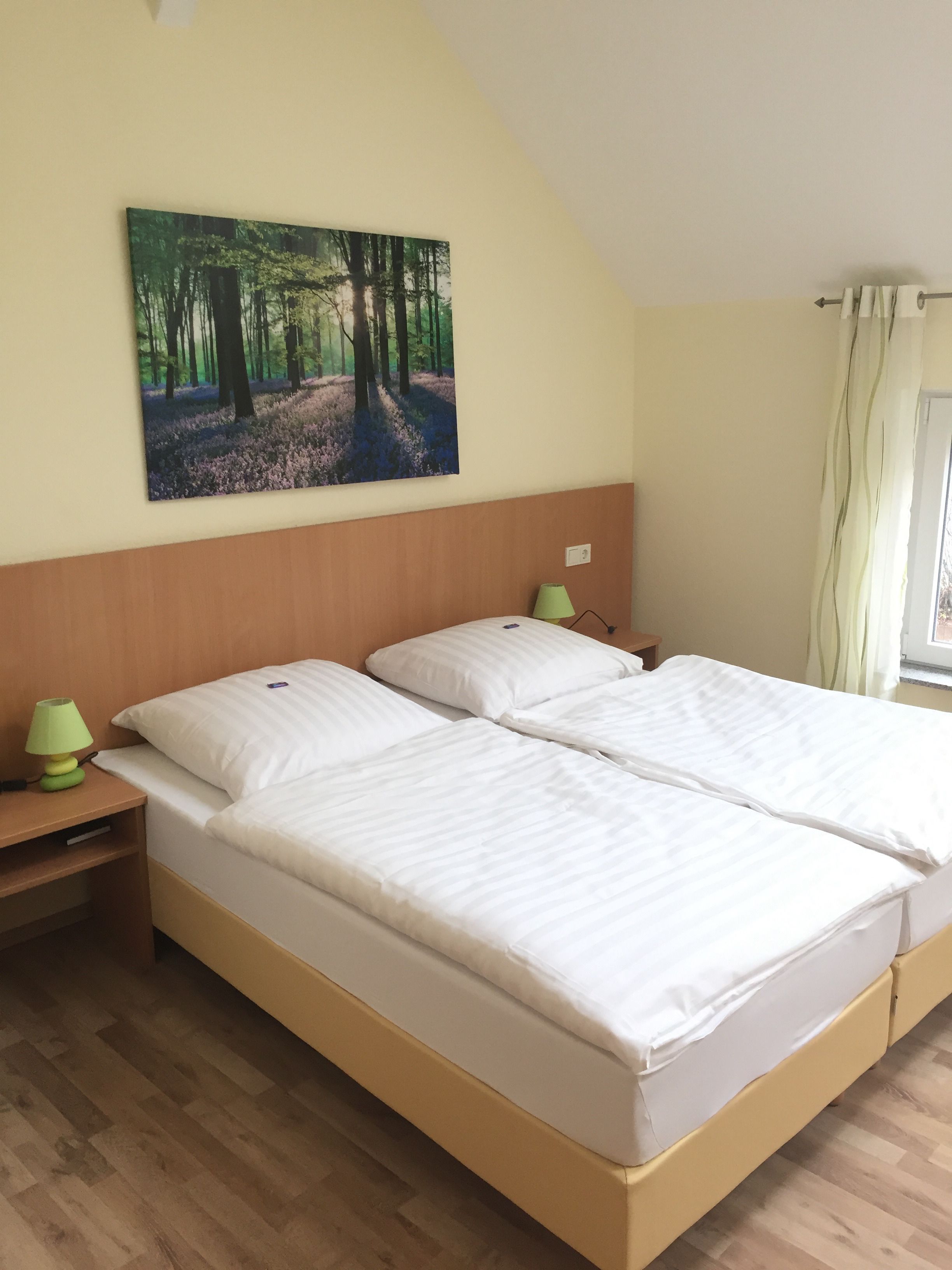 Uma cama de casal com roupa de cama branca, uma luminária verde e um quadro de uma floresta na parede.