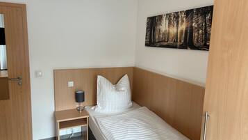 Um quarto pequeno e bem arrumado com uma cama de solteiro, uma mesa de cabeceira, uma luminária e um quadro com uma paisagem florestal na parede.