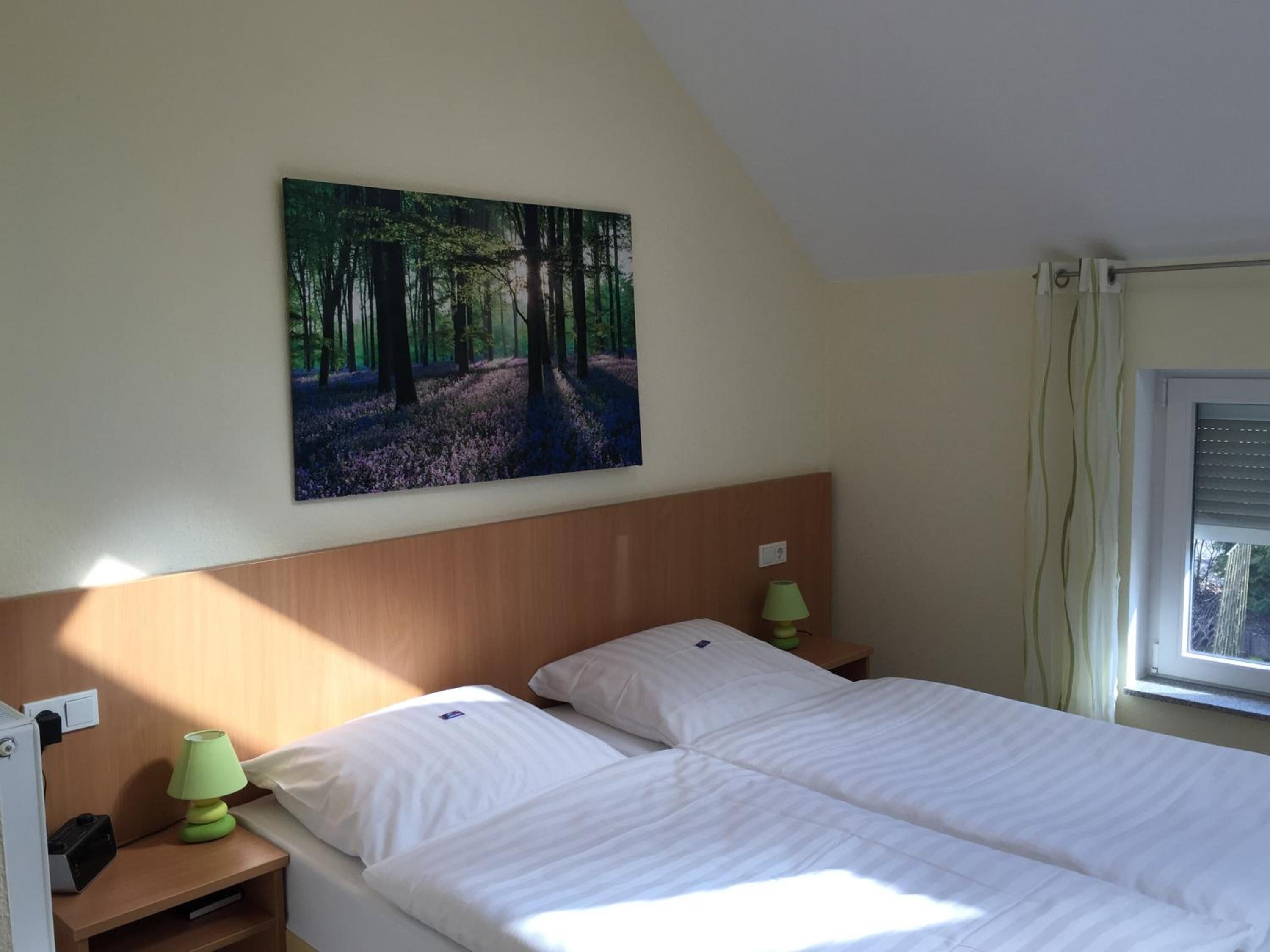 Quarto com duas camas, um quadro de uma floresta na parede, uma janela com cortinas e uma mesa de cabeceira com abajur.