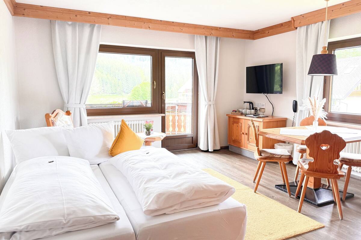 Quarto com cama, escrivaninha e cadeira. Há uma janela com cortinas, televisão na parede e teto de madeira.