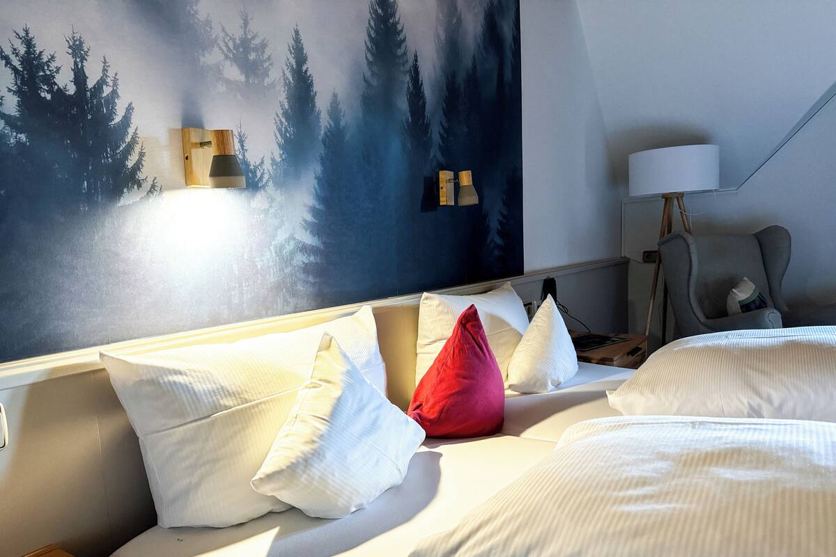 Quarto de hotel com uma cama, almofadas, uma luminária e uma parede com um mural de floresta.