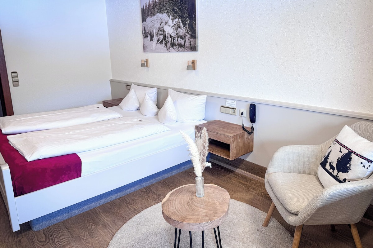 Quarto de hotel com cama, mesa de cabeceira, cadeira e telefone fixado na parede.