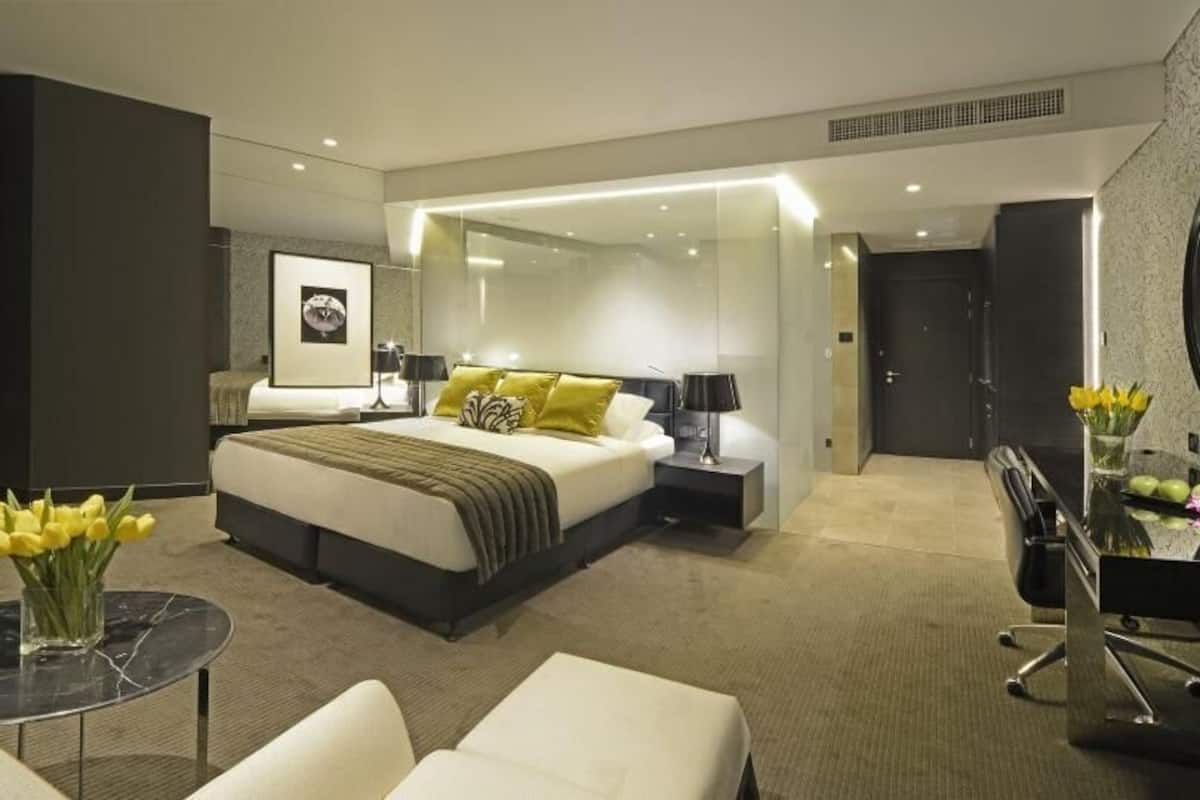 Quarto de hotel moderno com uma cama grande, mesas de cabeceira, escrivaninha, cadeira, espelho redondo e um vaso com flores.