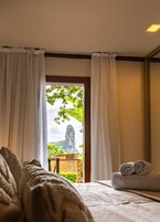 Quarto com uma cama, uma mesinha com uma toalha dobrada e vista para uma paisagem natural pela janela com cortinas.