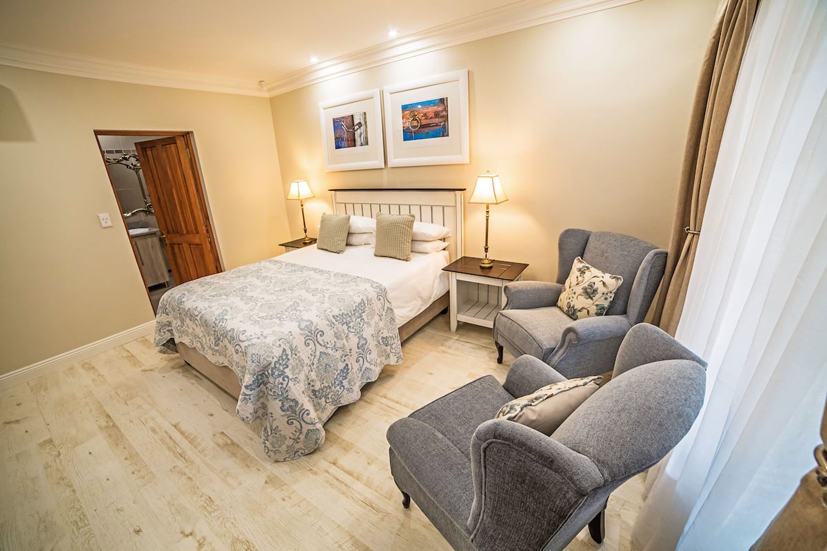 Quarto de hotel com uma cama, duas poltronas, uma mesa de cabeceira e uma porta de madeira.