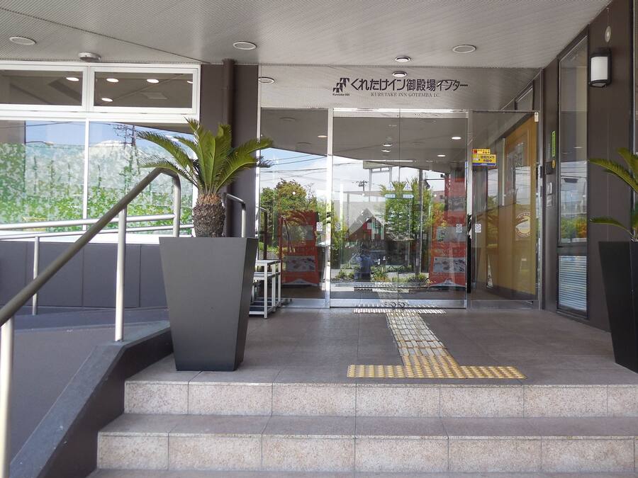 Entrada de hotel com rampa para a porta, vasos de plantas e fachada de vidro transparente.
