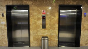 Dois elevadores com visor digital e uma lixeira num hall de entrada com paredes de mármore.
