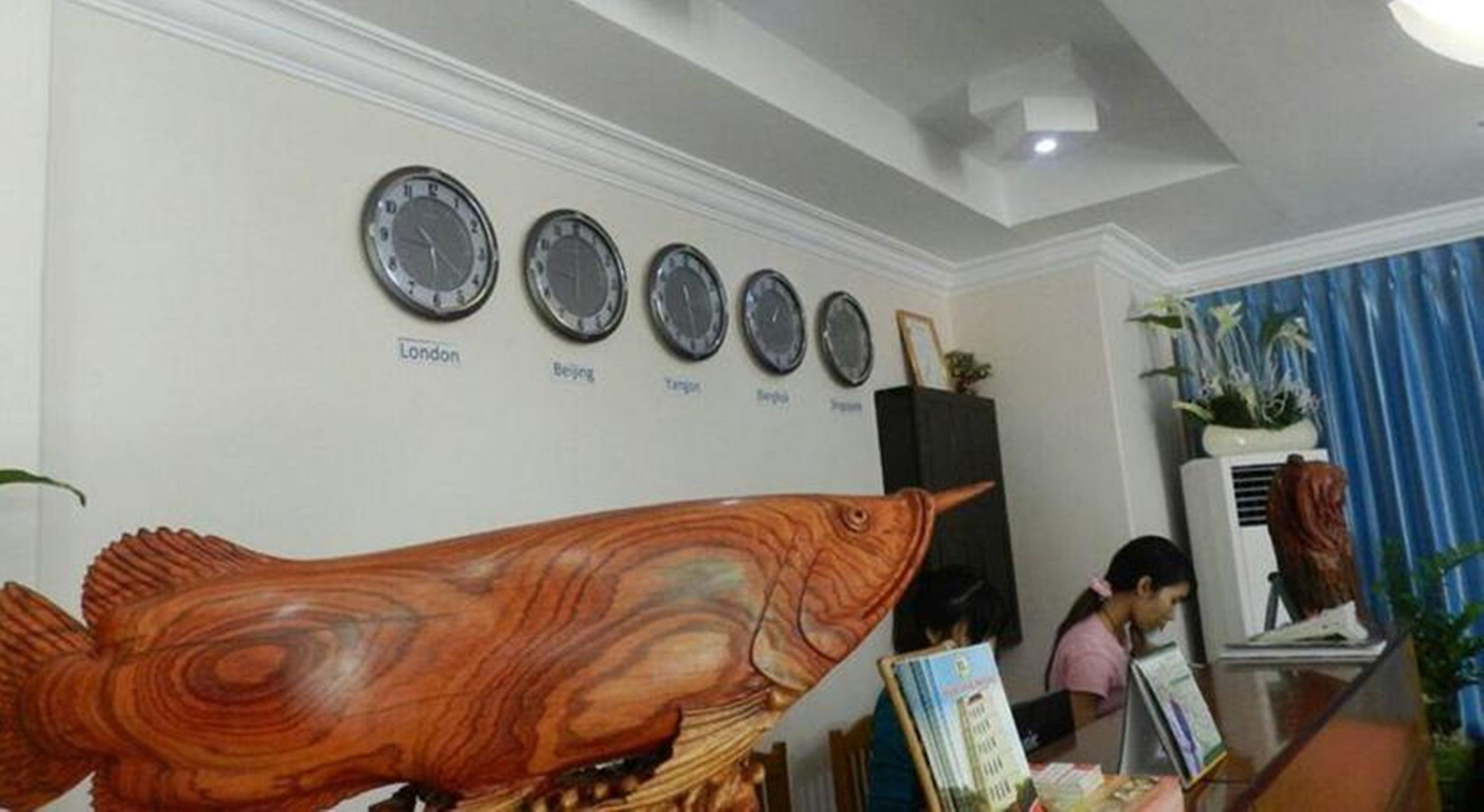 Quarto com uma grande escultura de peixe de madeira, um relógio de parede com a inscrição 'London' e uma pessoa trabalhando em uma mesa.