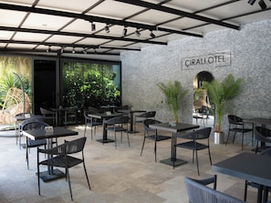 Uma área moderna de café ao ar livre, com mesas e cadeiras, um letreiro com a inscrição 'CIRALI HOTEL' e plantas em vasos.