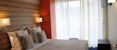 Quarto de hotel com uma cama grande, televisão na parede e janela com cortinas leves.