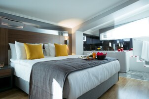 Quarto de hotel moderno com uma cama grande, uma mesa de cabeceira, uma pequena escrivaninha e um banheiro visível através de um painel de vidro.