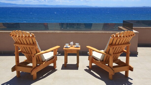 Duas cadeiras Adirondack de madeira com almofadas, uma mesa pequena com xícaras e um bule, e vista para o oceano.
