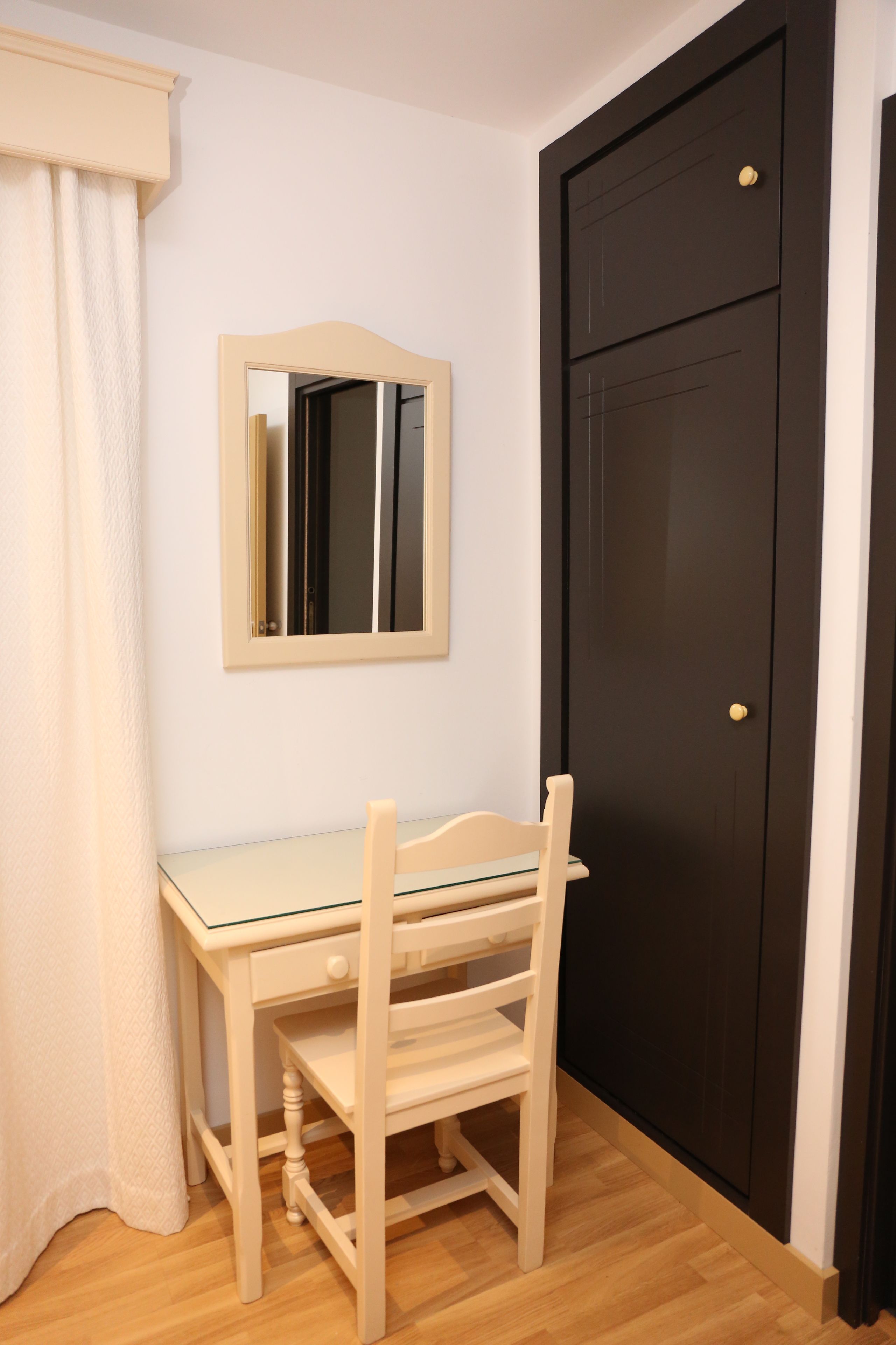 Um quarto pequeno com uma escrivaninha e cadeira de madeira, um espelho e uma porta escura.