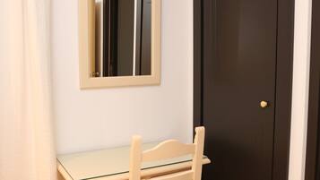Um quarto pequeno com uma escrivaninha e cadeira de madeira, um espelho e uma porta escura.