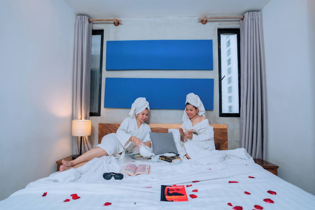 Quarto de hotel com uma cama, duas pessoas lendo, uma luminária e uma obra de arte azul na parede.