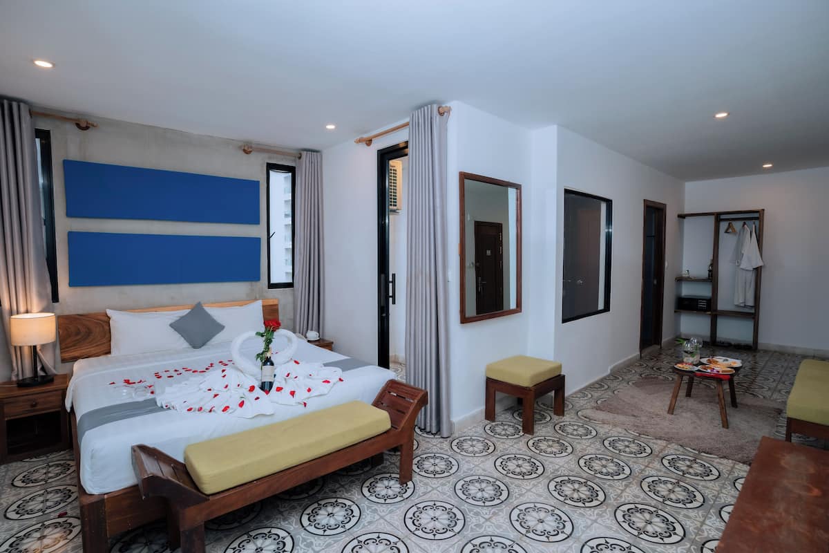 Quarto de hotel com uma cama grande, mesas de cabeceira, um espelho, um banquinho e um espaço de refeições com mesa e cadeiras.