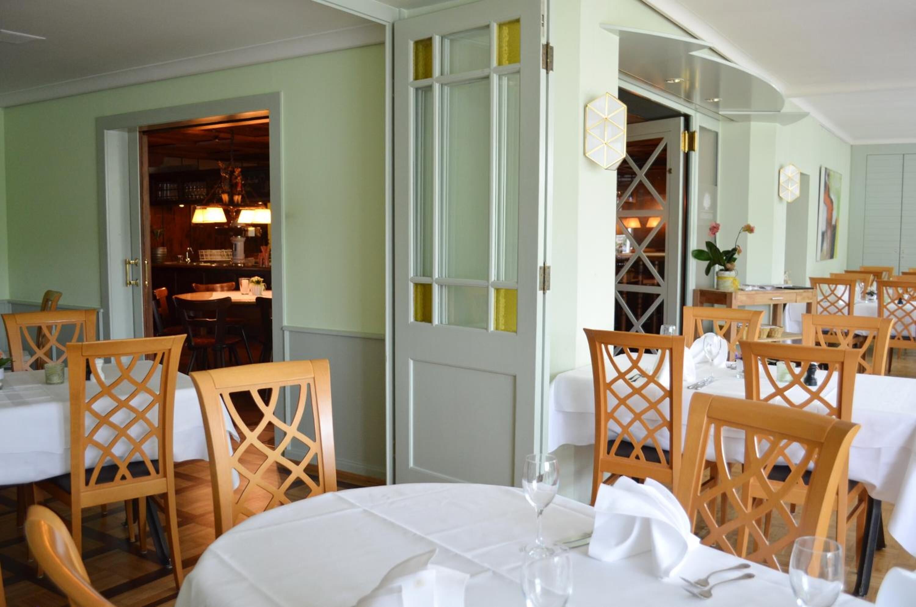 Uma área de refeições com mesas redondas e cadeiras de madeira, uma porta que dá para uma cozinha e um aplique na parede.