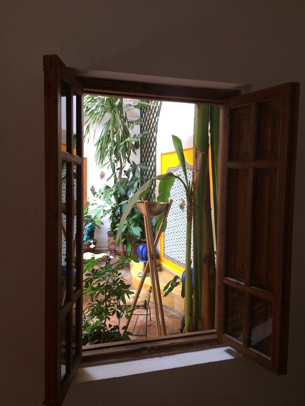 Quarto com uma janela grande que se abre para um pátio, com plantas em vasos e uma pequena mesa.