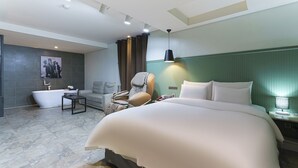 Quarto de hotel com uma cama grande, área de estar com sofá e poltrona, banheira e uma pequena mesa.