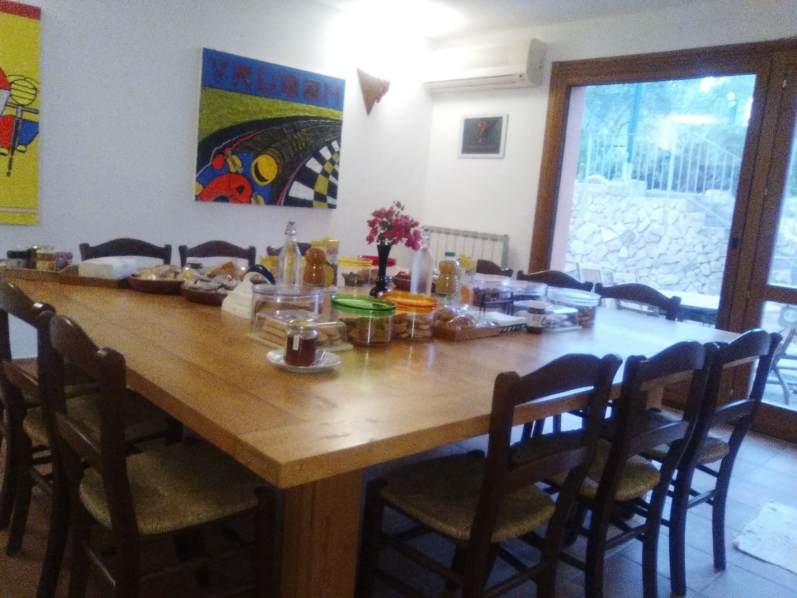 Uma sala de jantar com uma mesa de madeira comprida, cadeiras, um quadro e diversos alimentos.