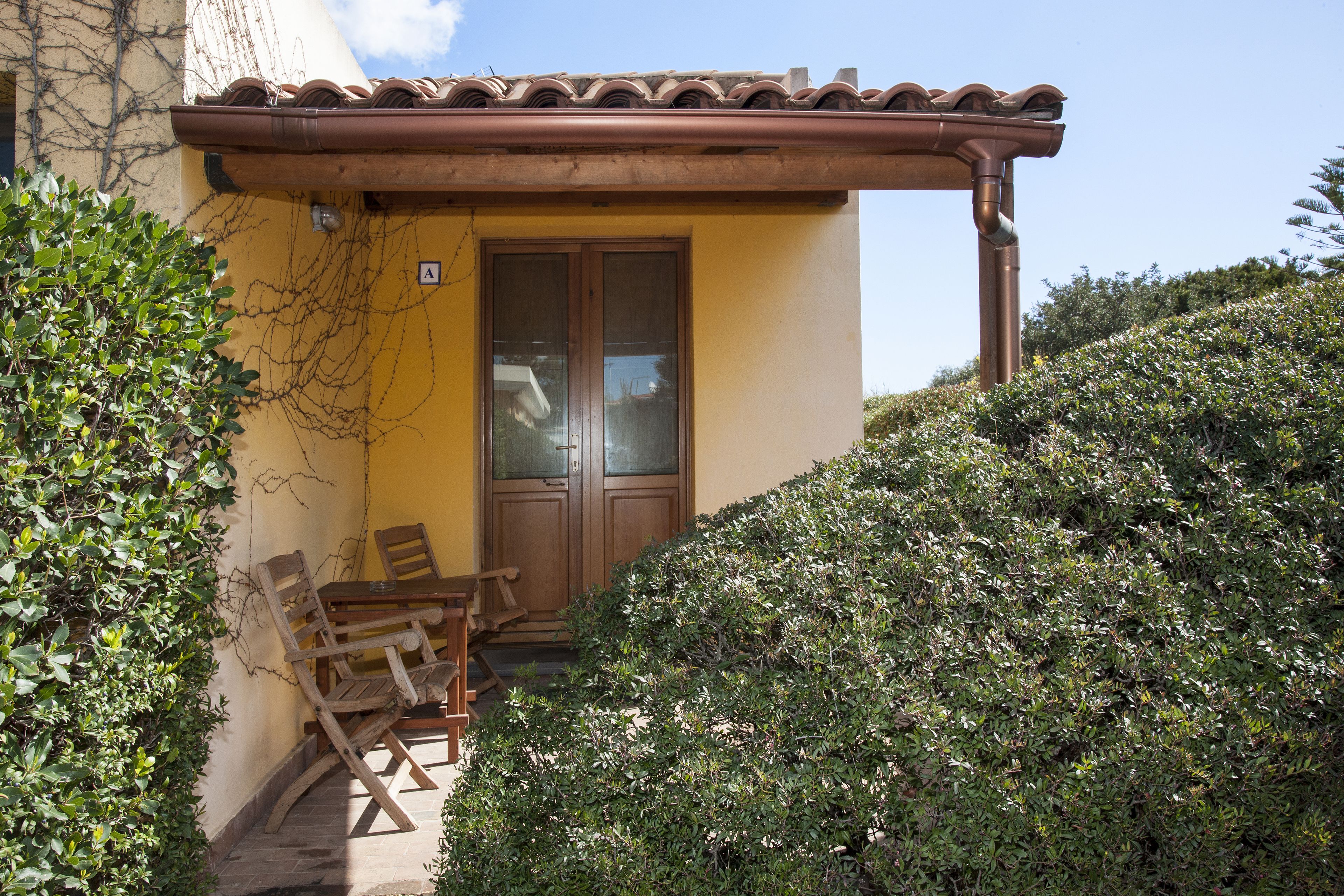 Um pequeno edifício amarelo com porta de madeira e telhado de telhas. Há um banco e uma mesa de madeira na parte externa, cercados por vegetação.