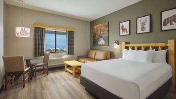 Quarto de hotel com uma cama grande, uma escrivaninha com duas cadeiras, um sofá e uma janela com vista. Há quadros nas paredes e uma luminária na escrivaninha.