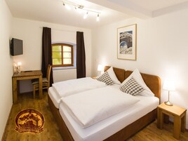 Quarto de hotel com cama de casal, escrivaninha, cadeira, janela com cortinas, televisão montada na parede e um quadro emoldurado.