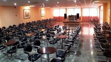 Uma sala de conferências com fileiras de cadeiras e mesas, um palco na frente e grandes janelas com cortinas.