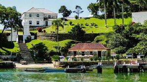 Uma paisagem costeira com um edifício branco, uma construção com telhado vermelho e barcos atracados na água.
