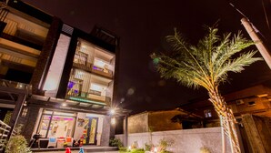 Um edifício moderno com entrada bem iluminada, varanda visível e área de jardim ajardinada com uma palmeira.