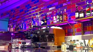 Uma área de bar com prateleiras de garrafas de bebidas, uma máquina de café e taças de vinho penduradas.