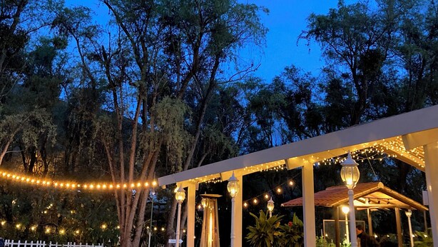 Uma área de refeições ao ar livre com luzes de corda, uma pérgola branca e uma cerca branca de piquete.