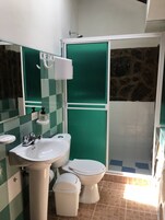 Casa de banho com lavatório, sanita e chuveiro. As paredes estão revestidas a azulejos verdes e brancos, e há uma janela com vista para o exterior.