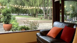 Quarto aconchegante com sofá de couro, mesa de madeira e uma grande janela com vista para um jardim com árvores e cerca branca.