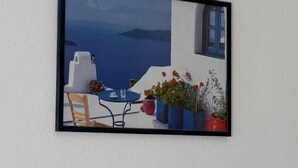 Pintura emoldurada retratando uma paisagem costeira, com uma cadeira, uma mesa e plantas em vasos.