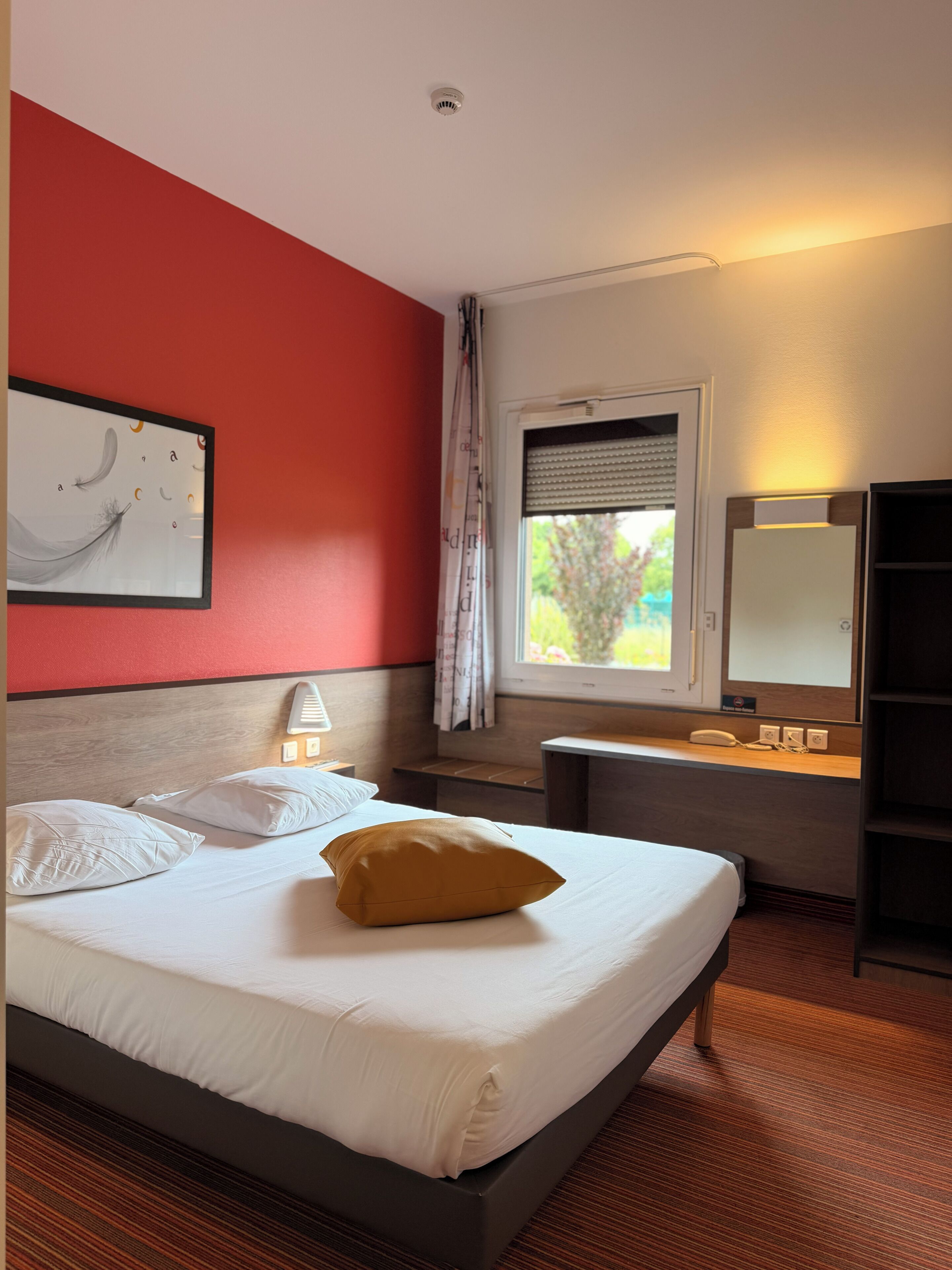 Quarto de hotel com cama, mesa de cabeceira, luminária, janela com persianas e espelho.