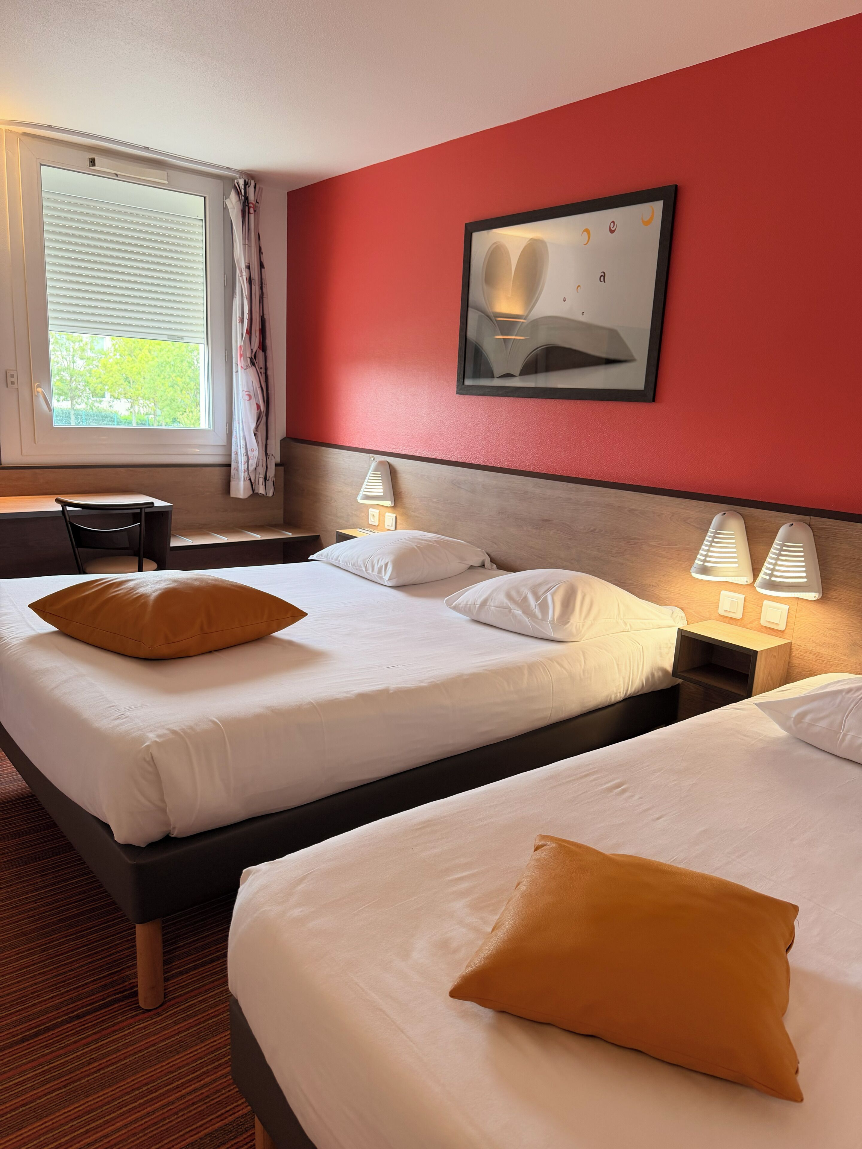 Quarto de hotel com duas camas, uma escrivaninha, uma cadeira, janela com cortinas e um quadro pendurado na parede.