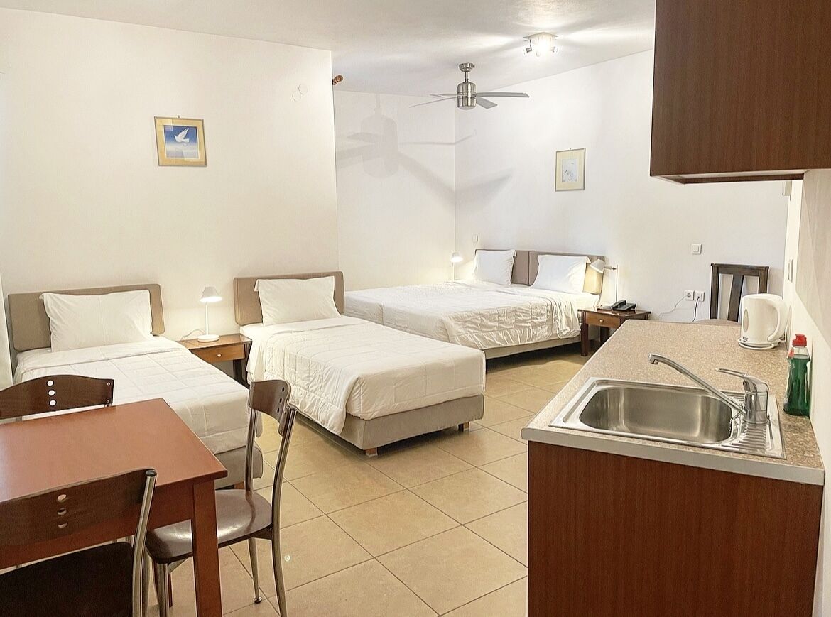 Quarto de hotel com duas camas, uma kitchenette pequena, uma mesa de jantar e um ventilador de teto.