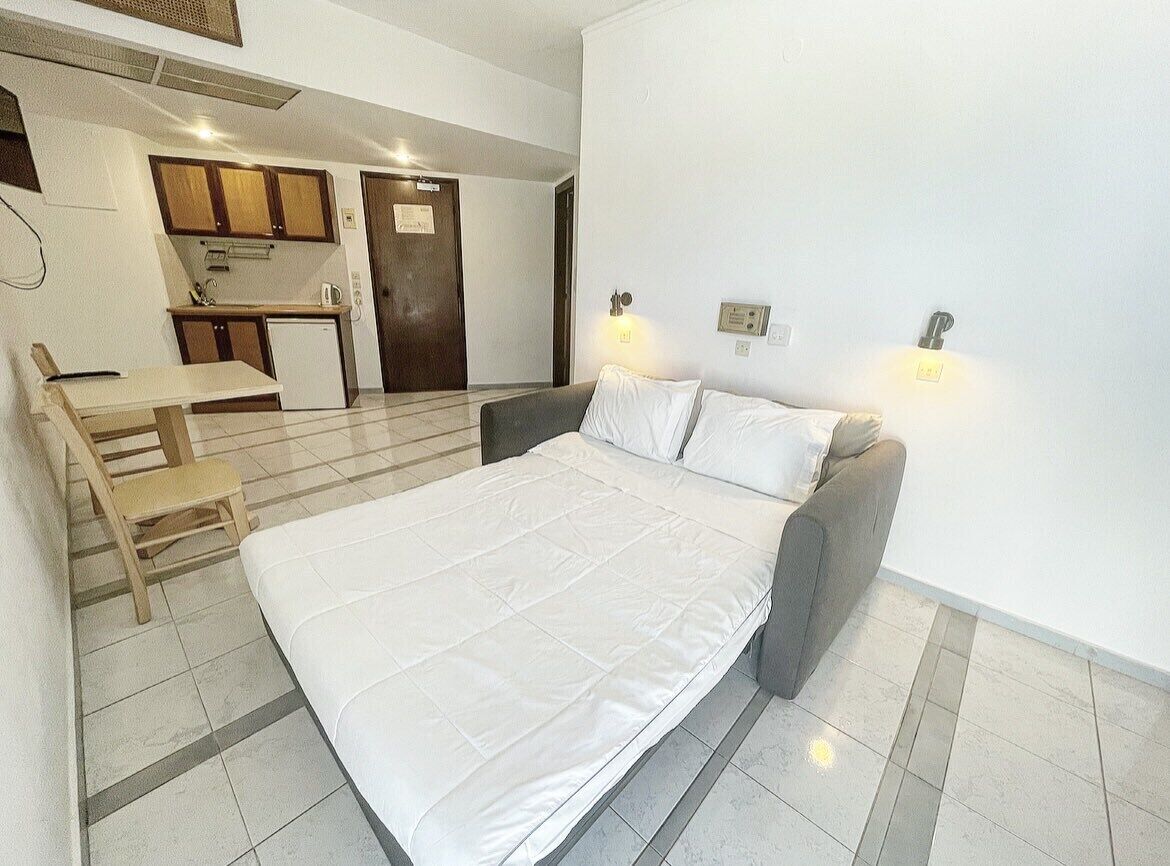 Quarto com cama, luminárias de cabeceira, uma pequena mesa de trabalho e uma kitchenette ao fundo.